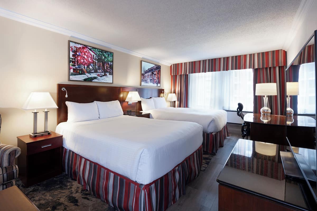 Quarto de hotel com duas camas, uma escrivaninha, uma cadeira, uma televisão e um quadro na parede.