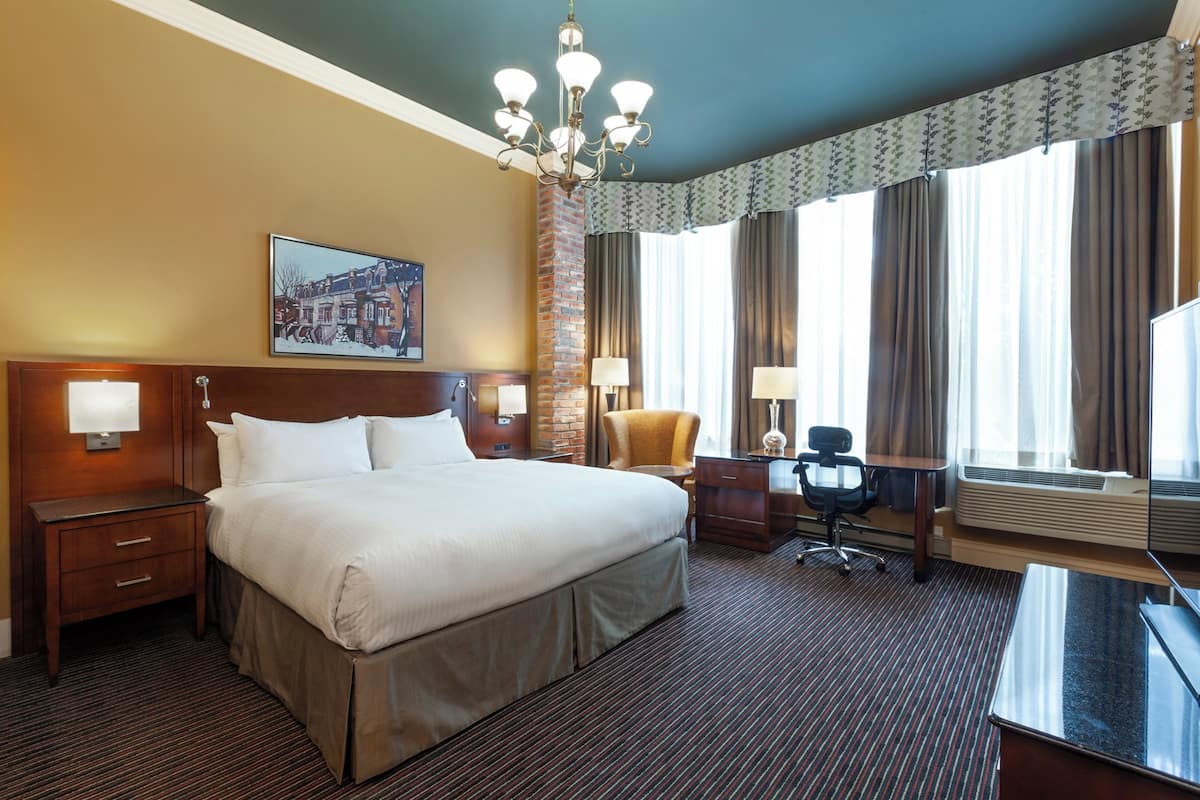 Quarto de hotel com uma cama grande, uma escrivaninha, uma cadeira, um abajur e um quadro na parede.