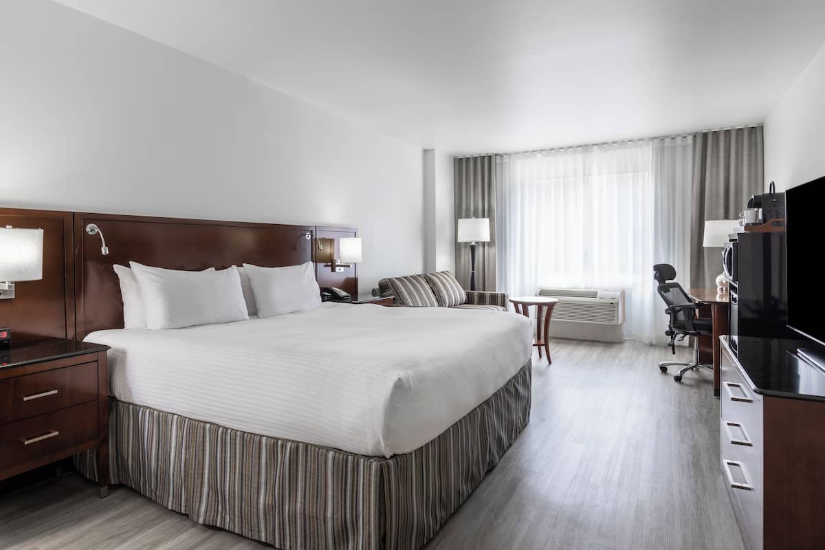 Quarto de hotel com uma cama grande, uma escrivaninha com cadeira, televisão e minibar.