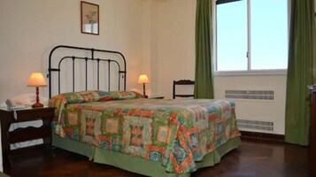 Quarto com cama, duas mesas de cabeceira com luminárias, uma mesinha de cabeceira menor, janela com cortinas e radiador.