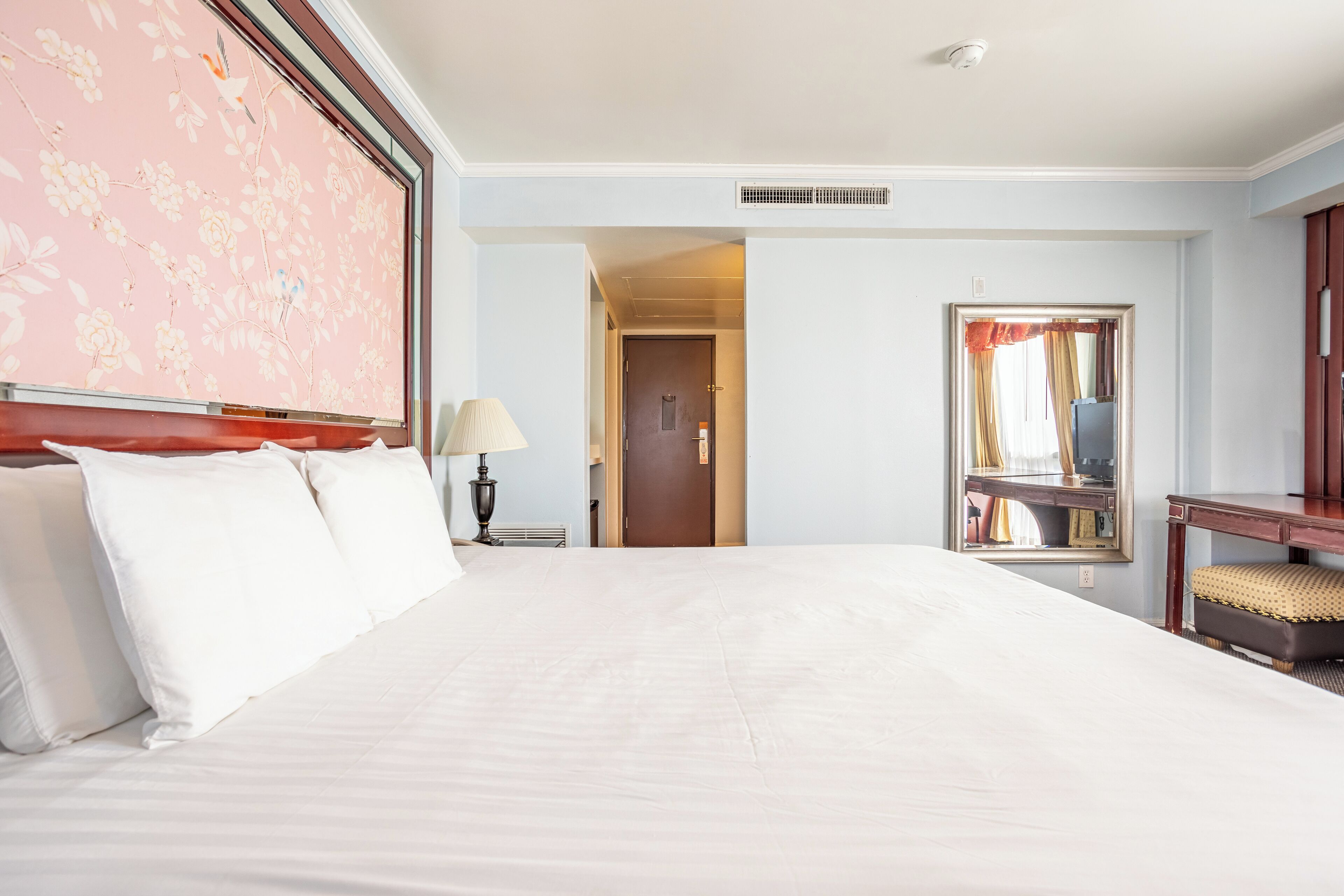 Quarto de hotel com uma cama grande, uma mesa com cadeira, um espelho e uma porta que dá para outro quarto.