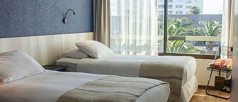 Quarto de hotel com duas camas, uma janela grande com cortinas, piso de madeira e vista para prédios e palmeiras.