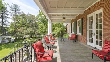Varanda coberta com cadeiras listradas de vermelho, uma mesa e vista para um jardim e árvores.