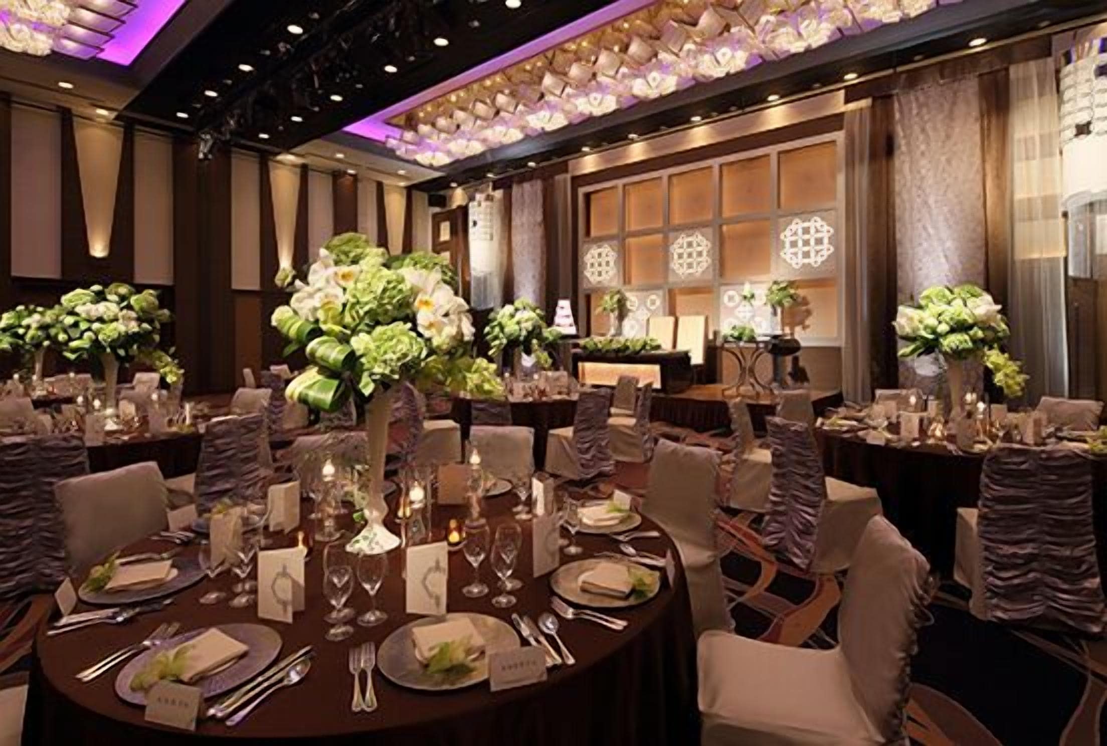 Uma sala de banquetes com mesas redondas preparadas para refeições, cada uma com louça posta e centros de mesa florais.