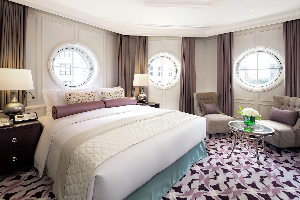 Quarto de hotel com uma cama grande, duas poltronas, uma pequena mesa com um vaso e duas janelas com cortinas.