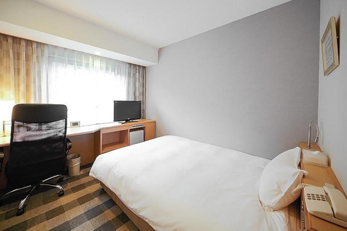 Quarto de hotel com cama, escrivaninha com computador, cadeira, televisão e telefone.