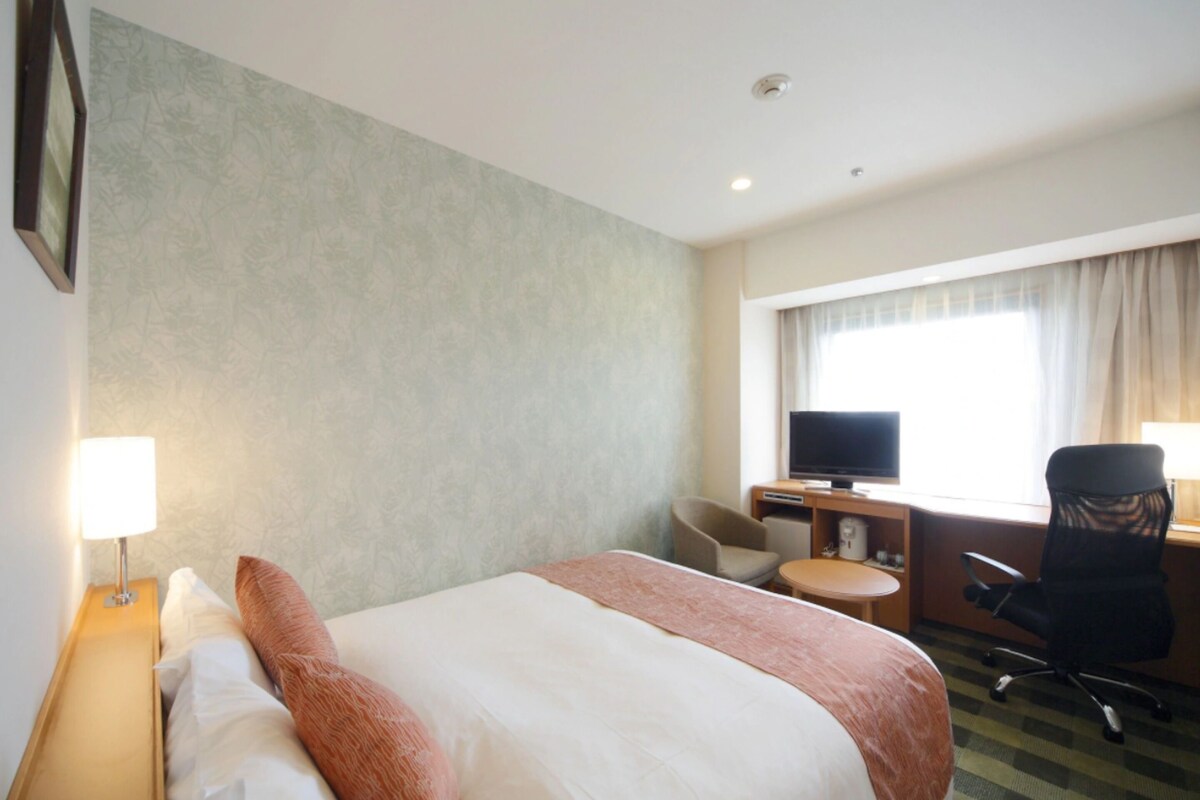 Quarto de hotel com cama, escrivaninha com televisão, cadeira e janela com cortinas.