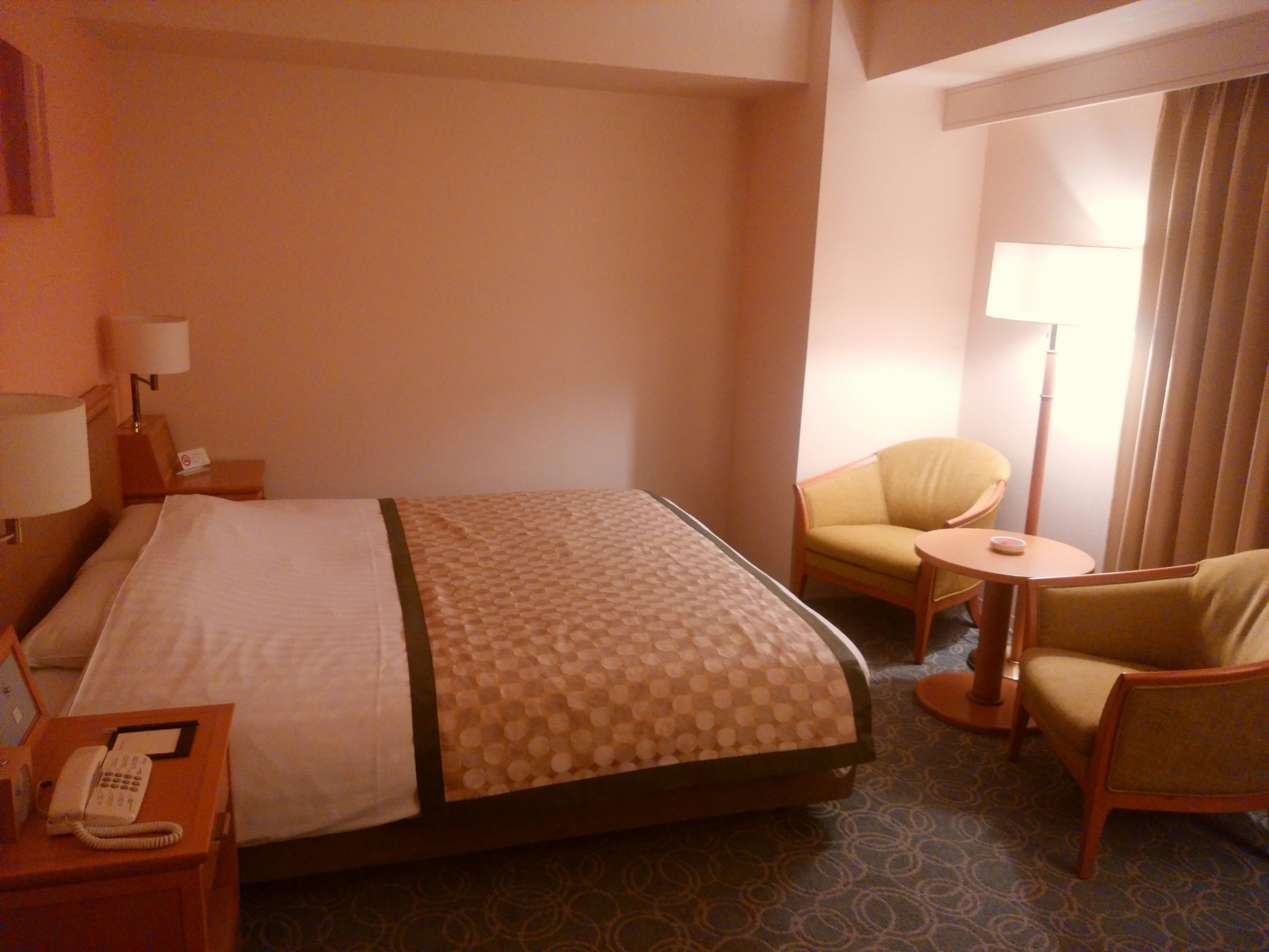 Quarto de hotel com cama, escrivaninha com telefone, cadeira, mesa pequena e abajur.