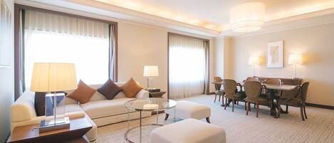 Quarto de hotel com sofá, apoio para os pés, mesa de centro de vidro, mesa de jantar com cadeiras, luminárias e um quadro pendurado na parede.