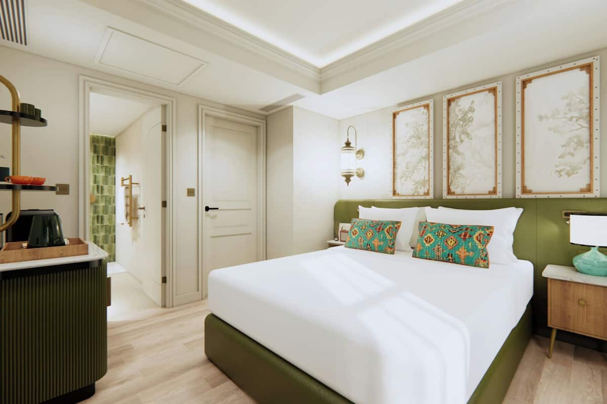 Quarto de hotel com uma cama grande, uma mesinha de cabeceira com luminária, um banco estofado verde e vista para um corredor com porta.