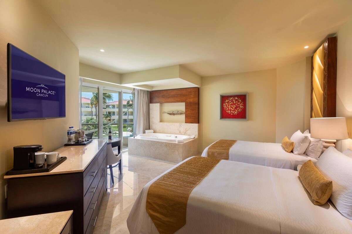 Quarto de hotel com duas camas, banheiro com banheira, escrivaninha com cafeteira e televisão exibindo o nome do hotel.