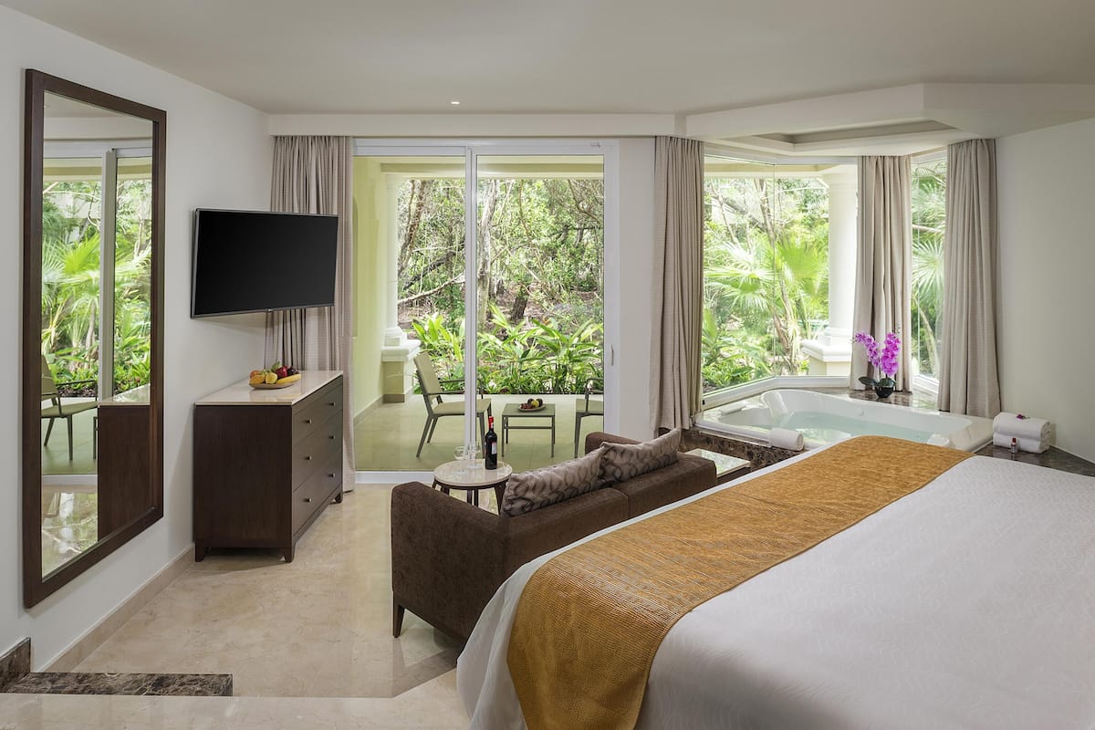 Quarto de hotel com uma cama grande, televisão, sofá, uma pequena mesa com uma garrafa, e vista para um jardim e piscina.