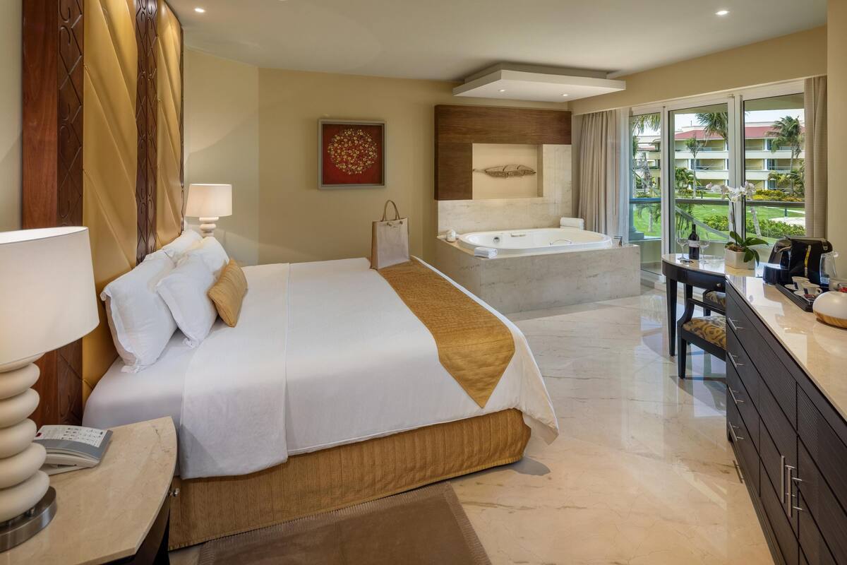 Quarto de hotel com uma cama grande, banheiro com banheira, escrivaninha com luminária e vista para o exterior.