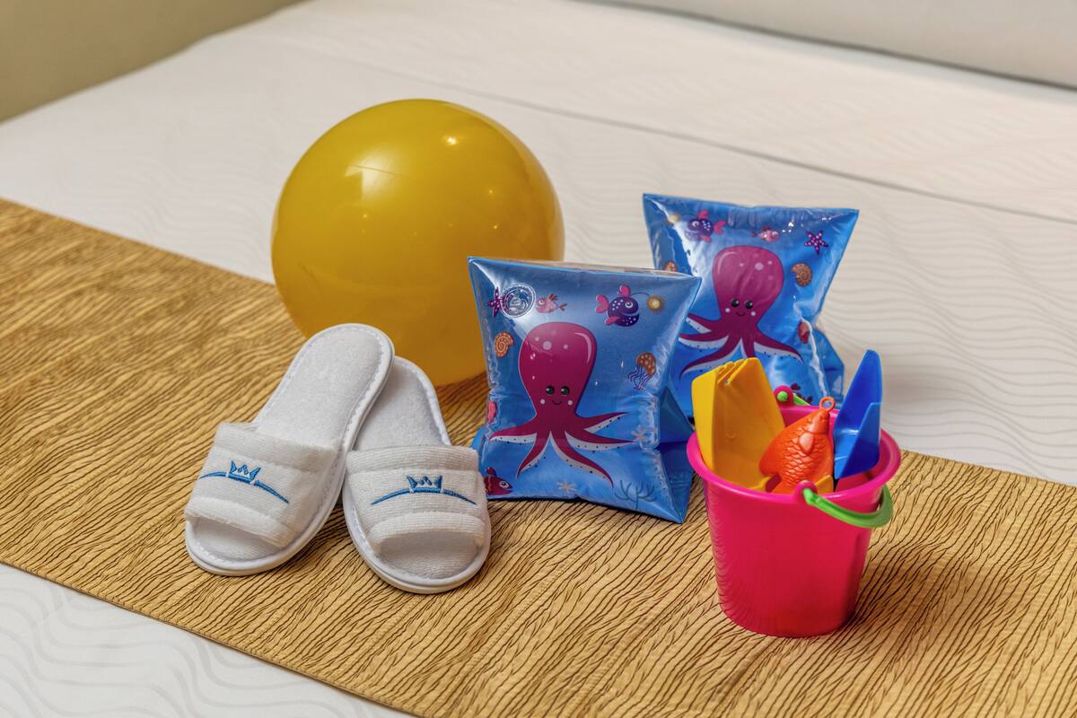 Cama com lençol branco, área com textura de madeira, bola amarela, chinelos brancos com coroa e sacolas azuis com estampas de polvo.