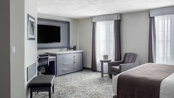 Quarto de hotel com cama, escrivaninha, cadeira, mesa pequena e televisão.