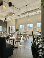 Uma sala de jantar moderna com mesas e cadeiras, iluminação pendente, janelas amplas e plantas em vasos.