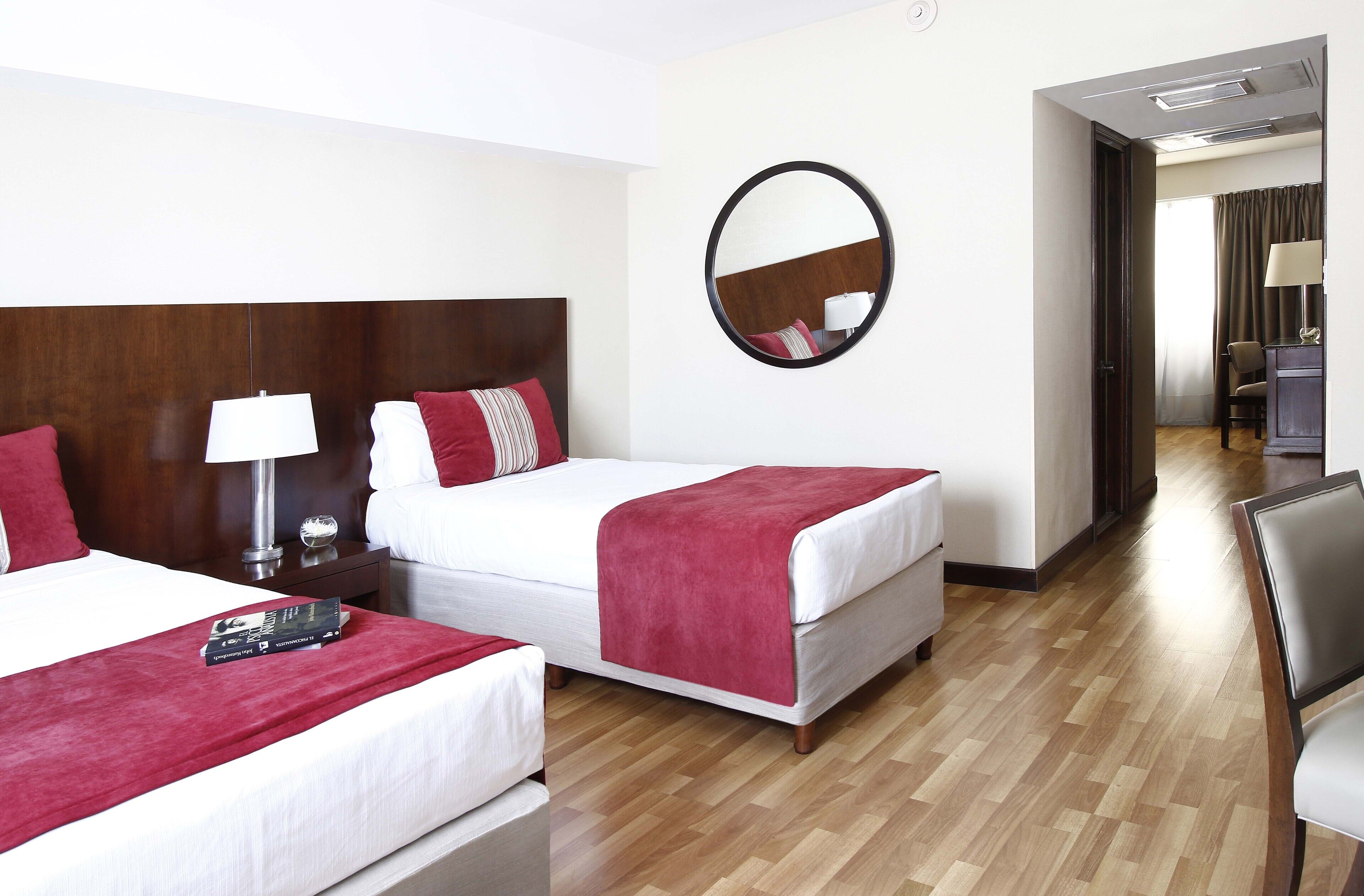 Quarto de hotel com duas camas, um espelho redondo, uma escrivaninha com cadeira e piso de madeira.