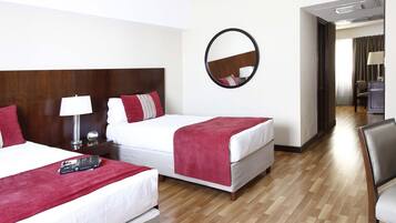 Quarto de hotel com duas camas, um espelho redondo, uma escrivaninha com cadeira e piso de madeira.