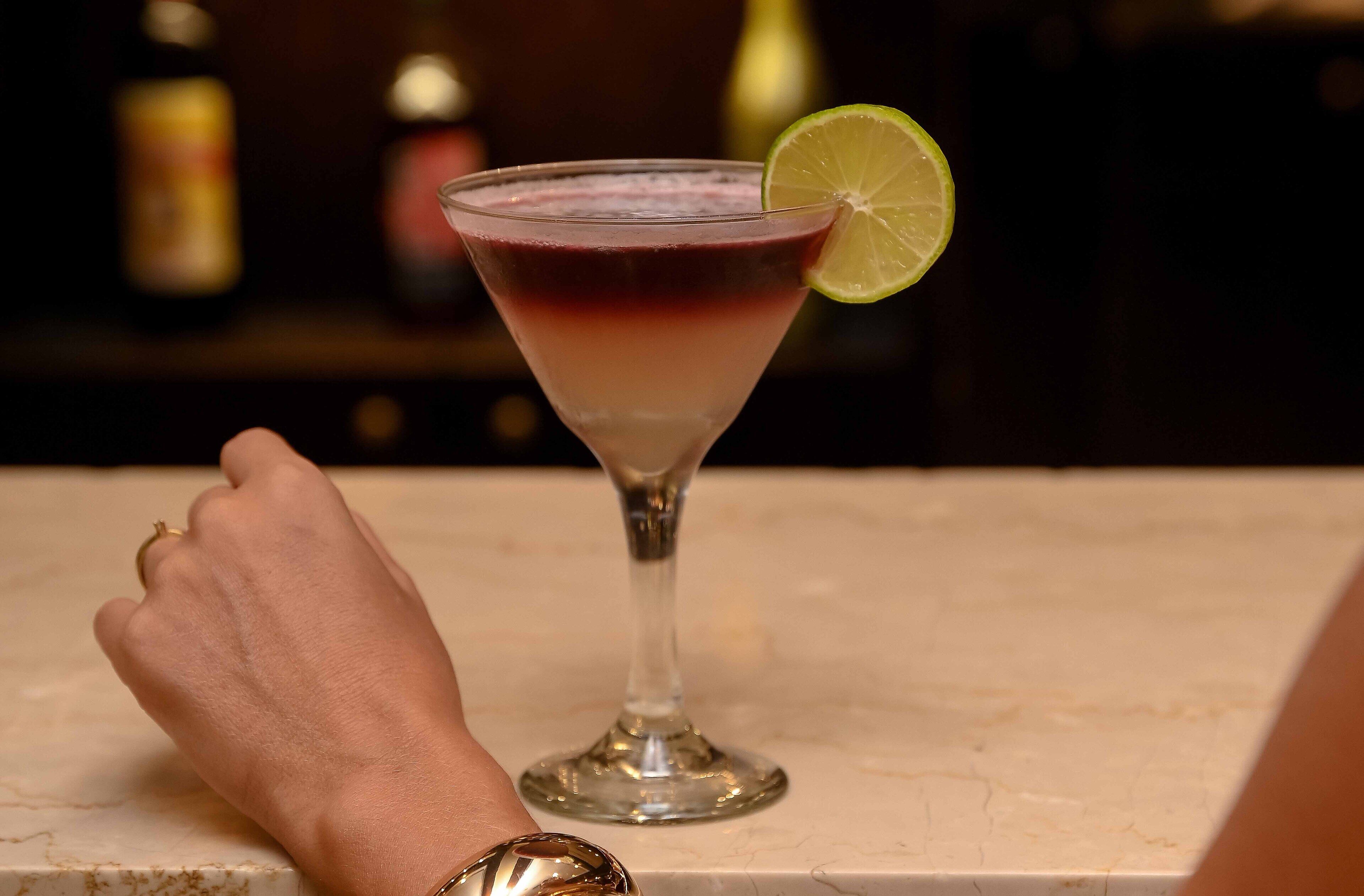 Um copo de martini com uma bebida em camadas e um toque de limão sobre uma superfície de mármore.