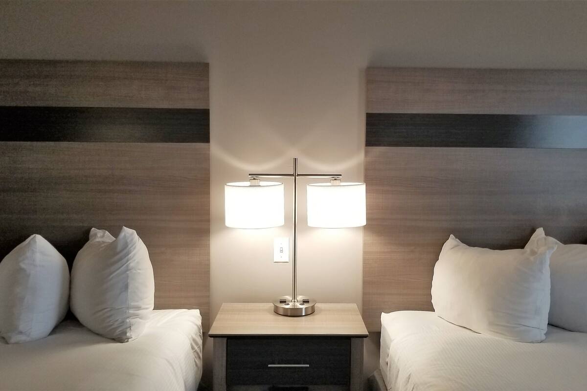 Quarto de hotel com duas camas, uma mesa de cabeceira com abajur e luminária na parede.