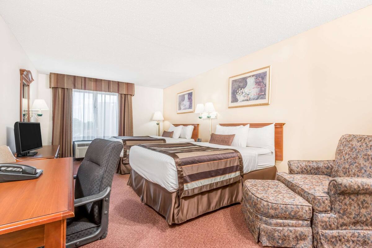 Quarto de hotel com duas camas, uma escrivaninha com computador, uma cadeira, um sofá e uma televisão.