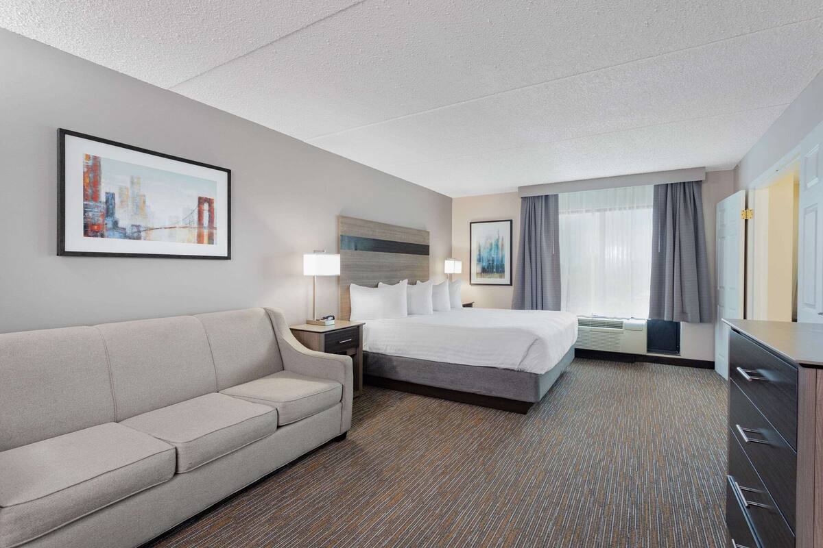 Quarto de hotel com cama, sofá, escrivaninha com luminária, cômoda e duas imagens emolduradas nas paredes.
