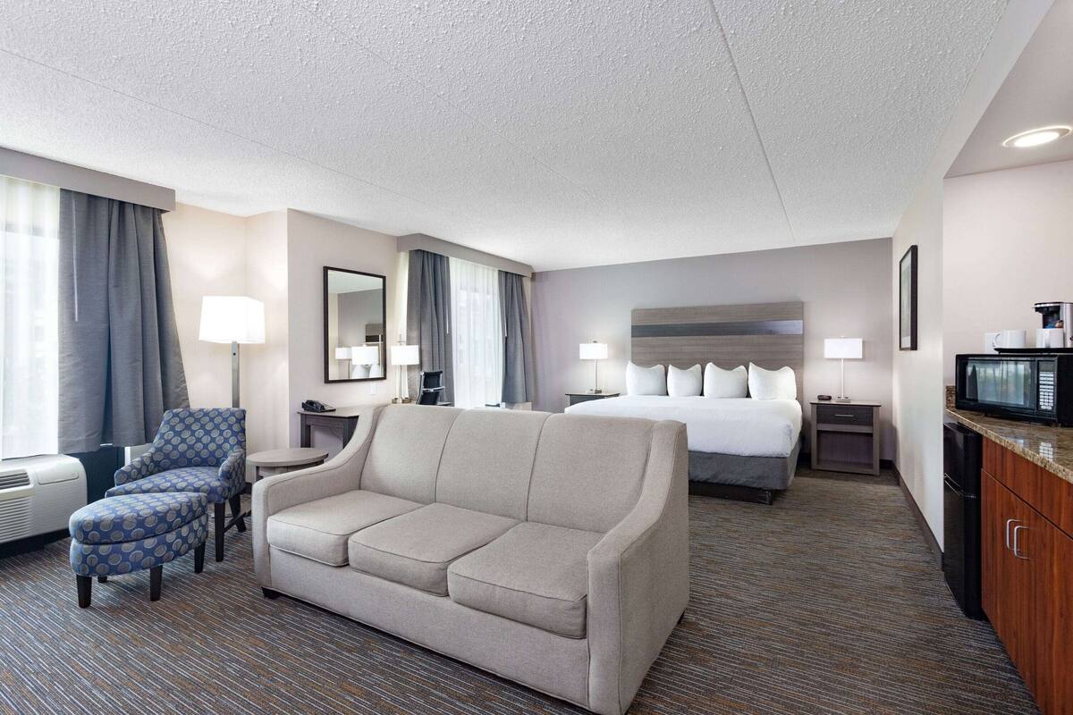Quarto de hotel com cama, sofá, cadeira, escrivaninha e cozinha compacta.