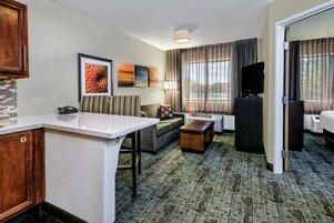 Quarto de hotel com cozinha compacta, sofá, mesa de centro, televisão e uma janela grande com cortinas.