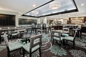 Uma sala de jantar de hotel com várias mesas e cadeiras, uma área de bar com parede de pedra ao fundo e iluminação no teto.