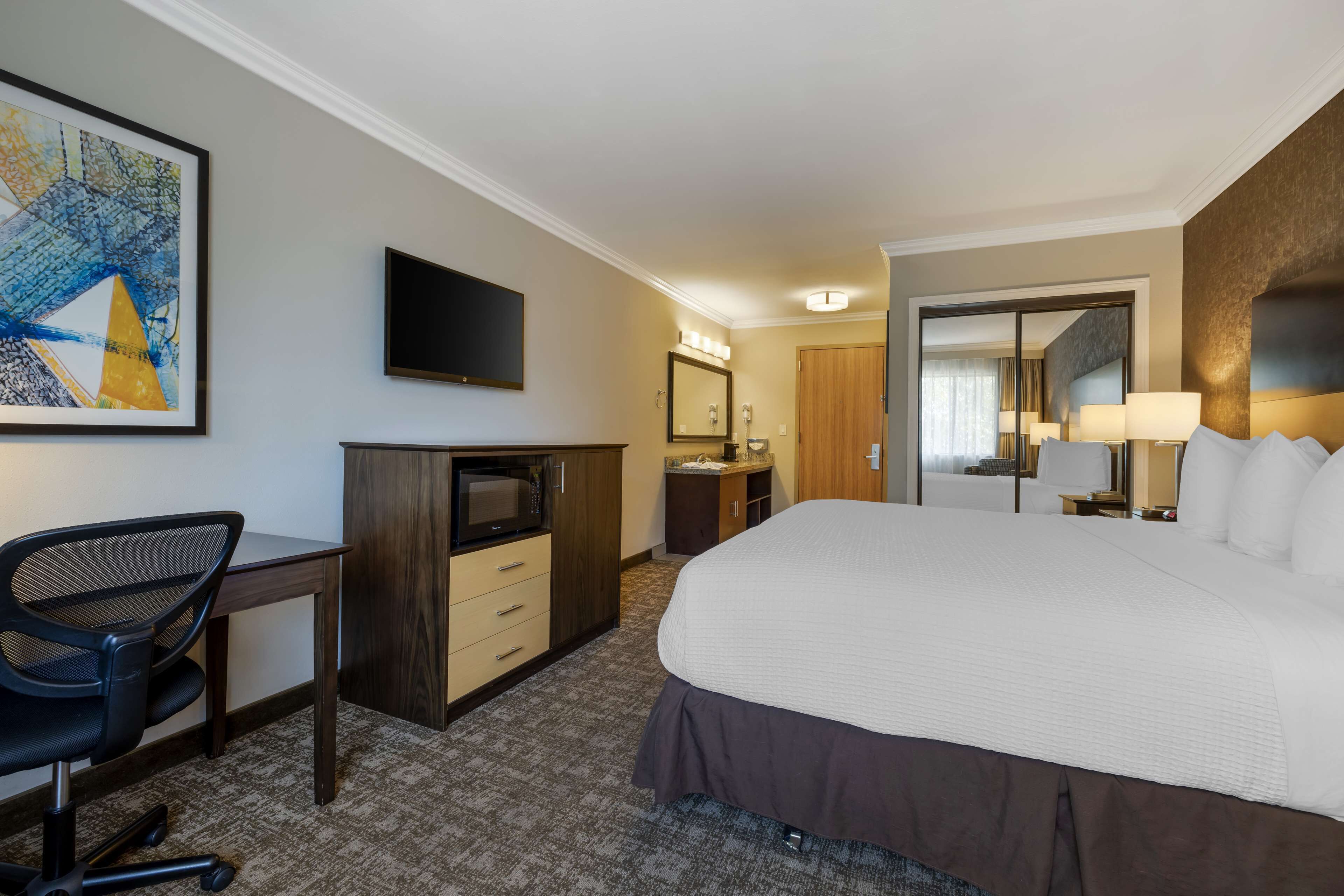 Quarto de hotel com uma cama grande, uma escrivaninha com cadeira, televisão, espelho e porta que dá para outro espaço.