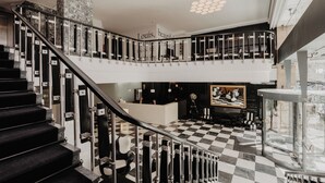 Um saguão de hotel com piso xadrez, balcão de recepção e escada com guarda-corpo de vidro.