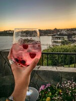 Uma mão segurando um copo com bebida rosa, morango e cubos de gelo, com vista para um rio e horizonte urbano ao pôr do sol.