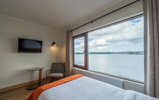 Quarto de hotel com cama, uma cadeira, uma mesa pequena, televisão e uma grande janela com vista para um corpo de água e terra distante.