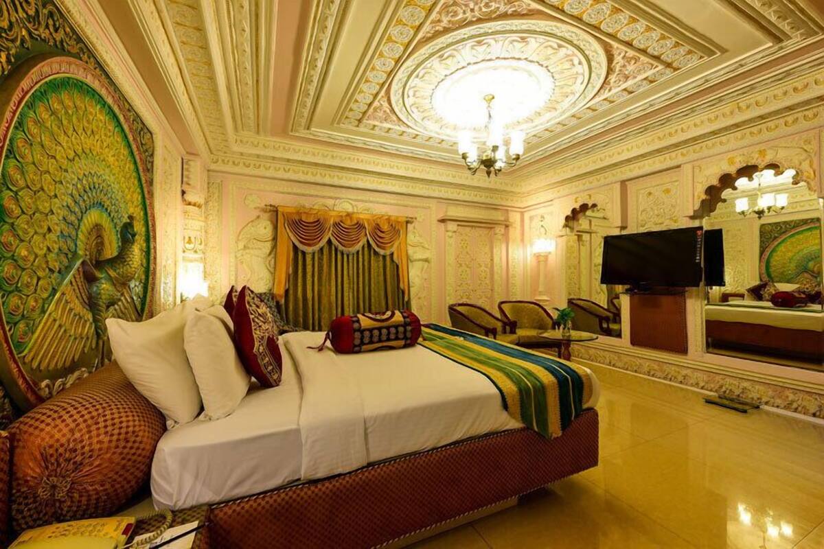 Quarto de hotel luxuoso com teto ornamentado, uma cama grande com várias almofadas, uma televisão e um espelho.