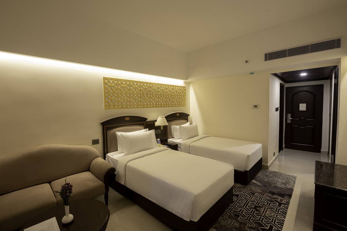 Quarto de hotel com duas camas, um sofá, um painel decorativo na parede e uma porta que dá para outro quarto.