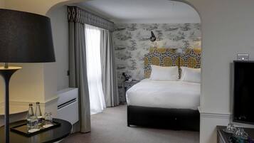 Quarto de hotel com cama, mesa de cabeceira, luminária, televisão e pequena mesa com copos e garrafas.
