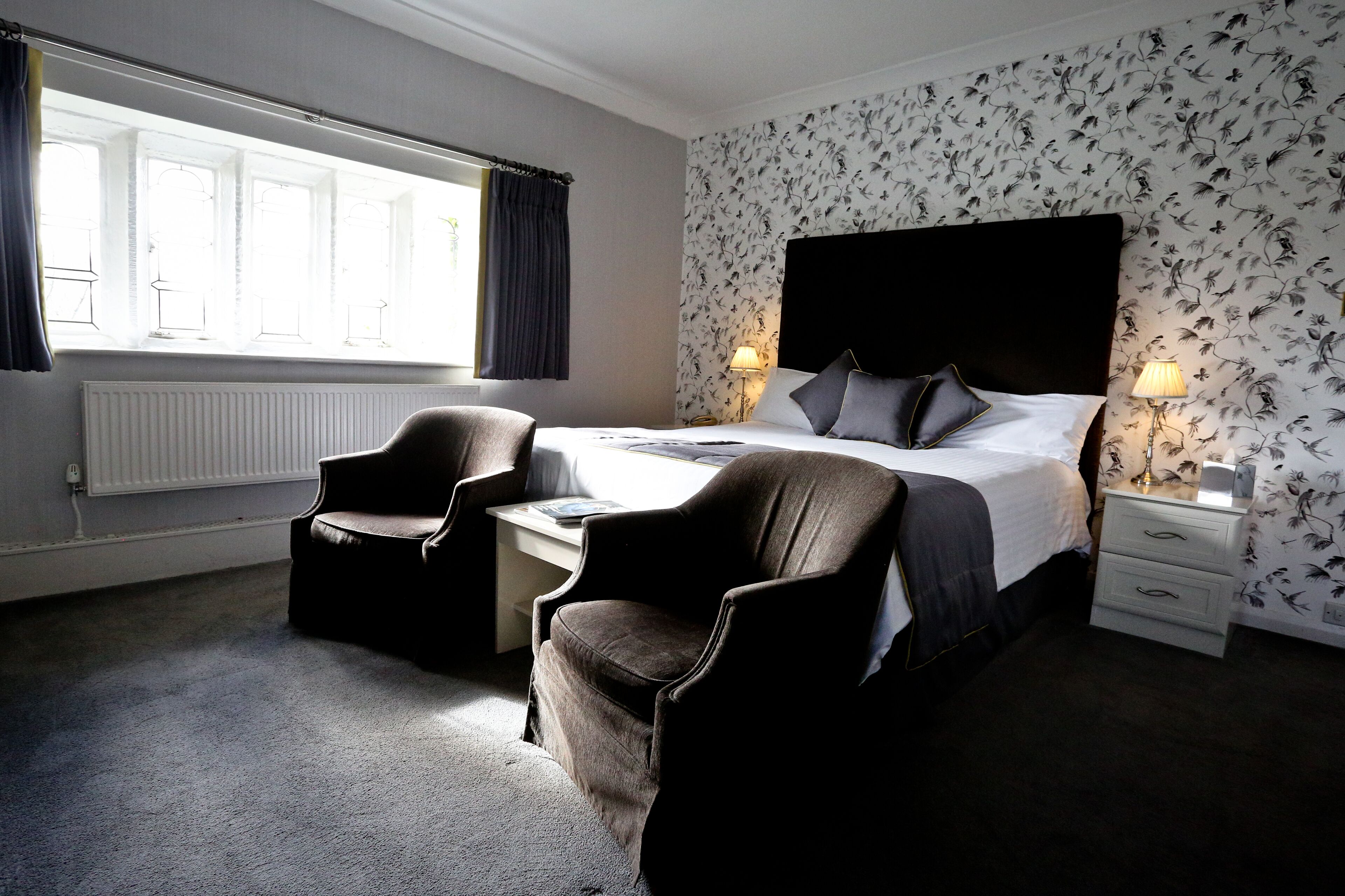 Quarto de hotel com uma cama grande, duas poltronas, uma mesa de trabalho e uma janela com cortinas.