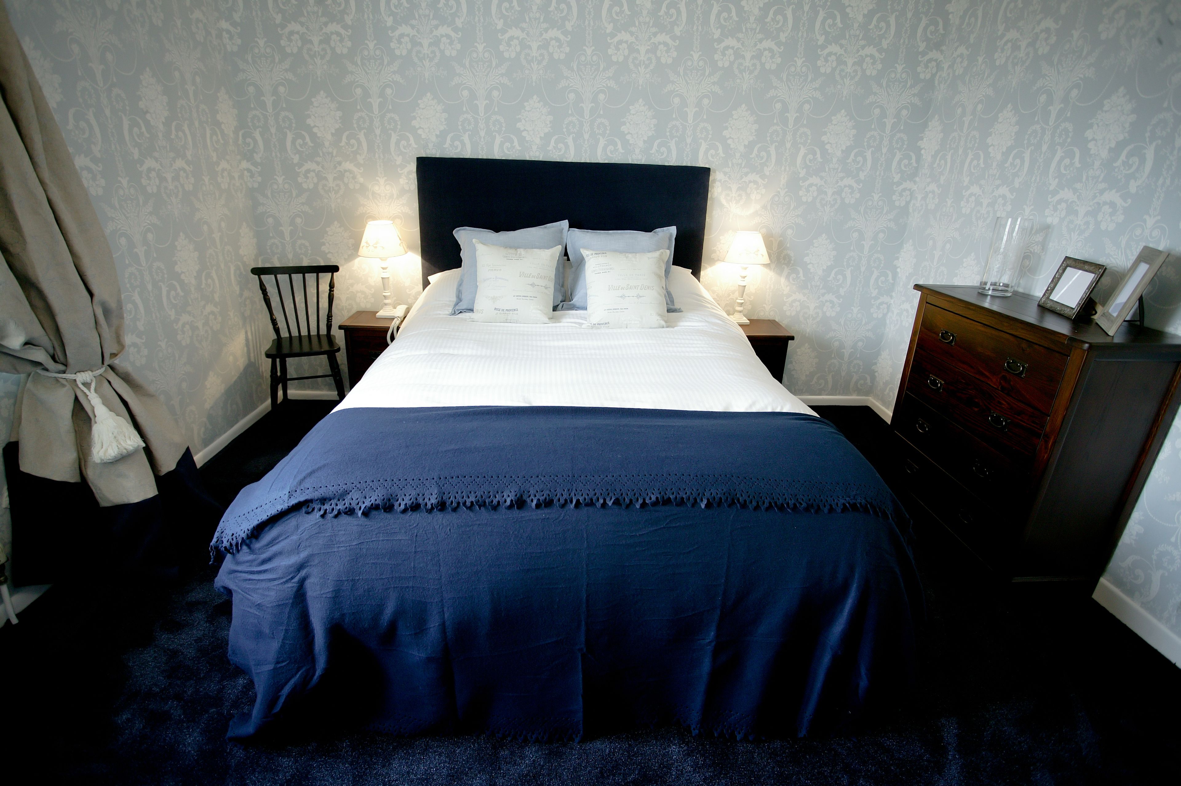 Cama bem arrumada com colcha azul escura e almofadas combinando, ladeada por duas mesas de cabeceira com abajures. Um aparador de madeira com espelho e moldura de quadro está à direita.