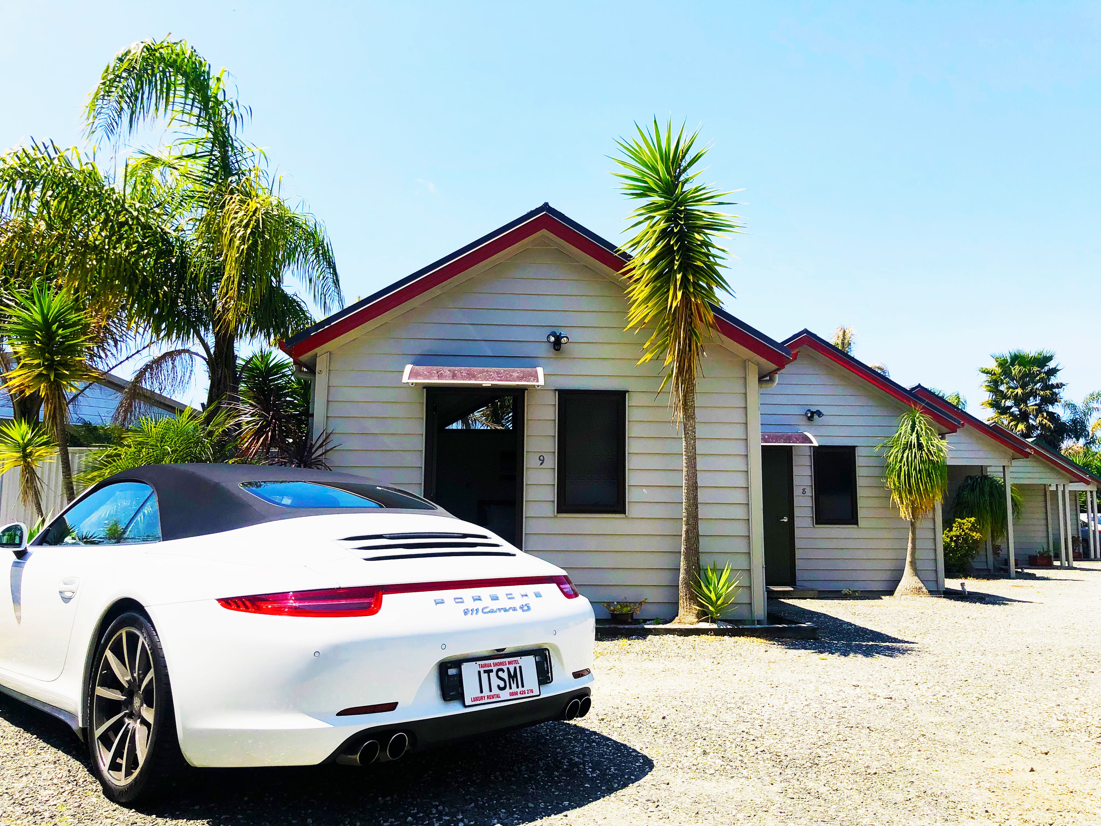 Um Porsche conversível branco estacionado em frente a um pequeno edifício térreo com telhado vermelho e uma placa que diz 'Tauris Shires Motel'.