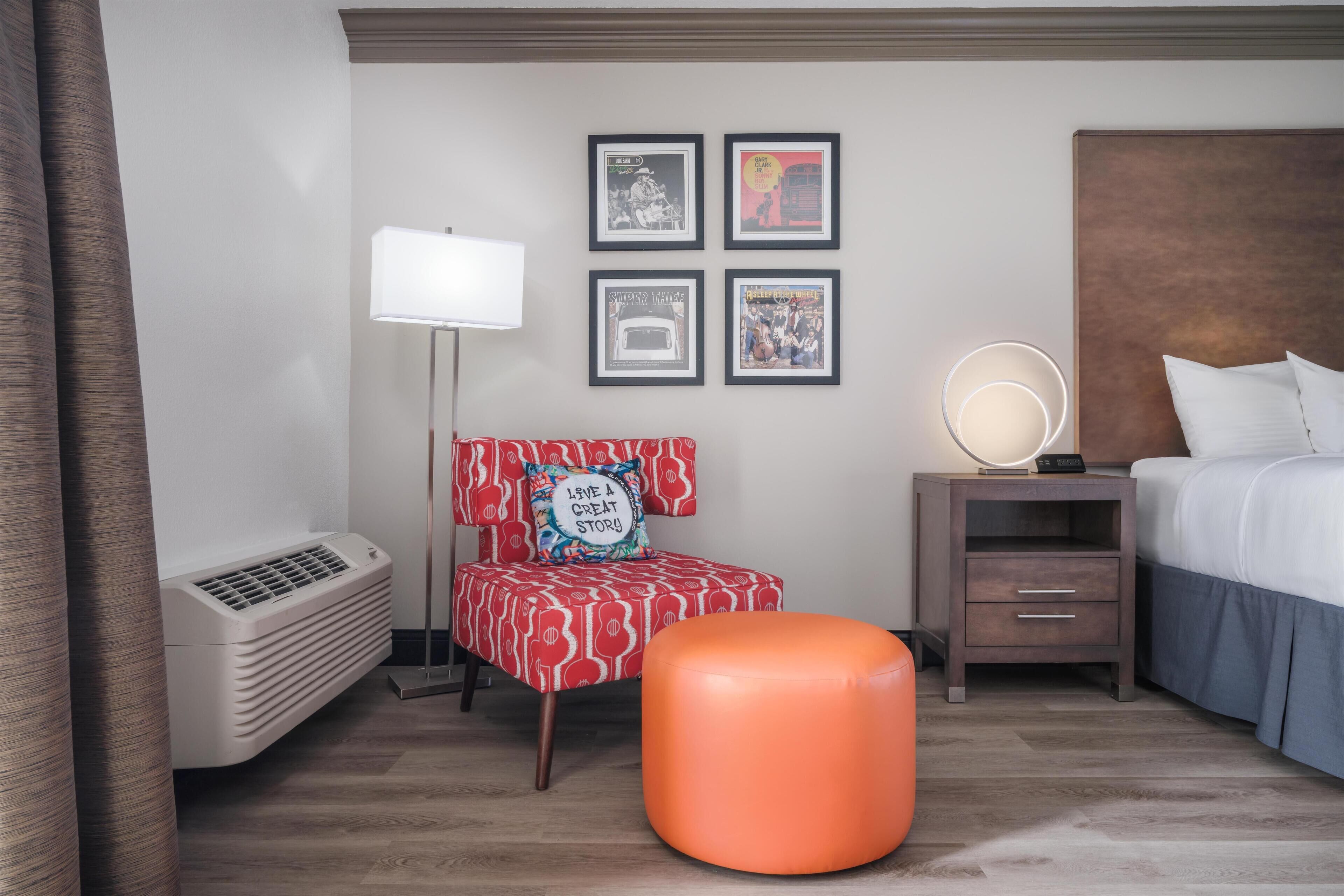 Quarto de hotel com cama, mesa de cabeceira com abajur, cadeira com almofada, aquecedor e quadros nas paredes.