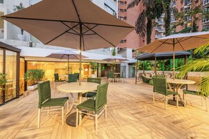 Um pátio externo com mesas redondas e cadeiras verdes, rodeado por grandes guarda-sóis.