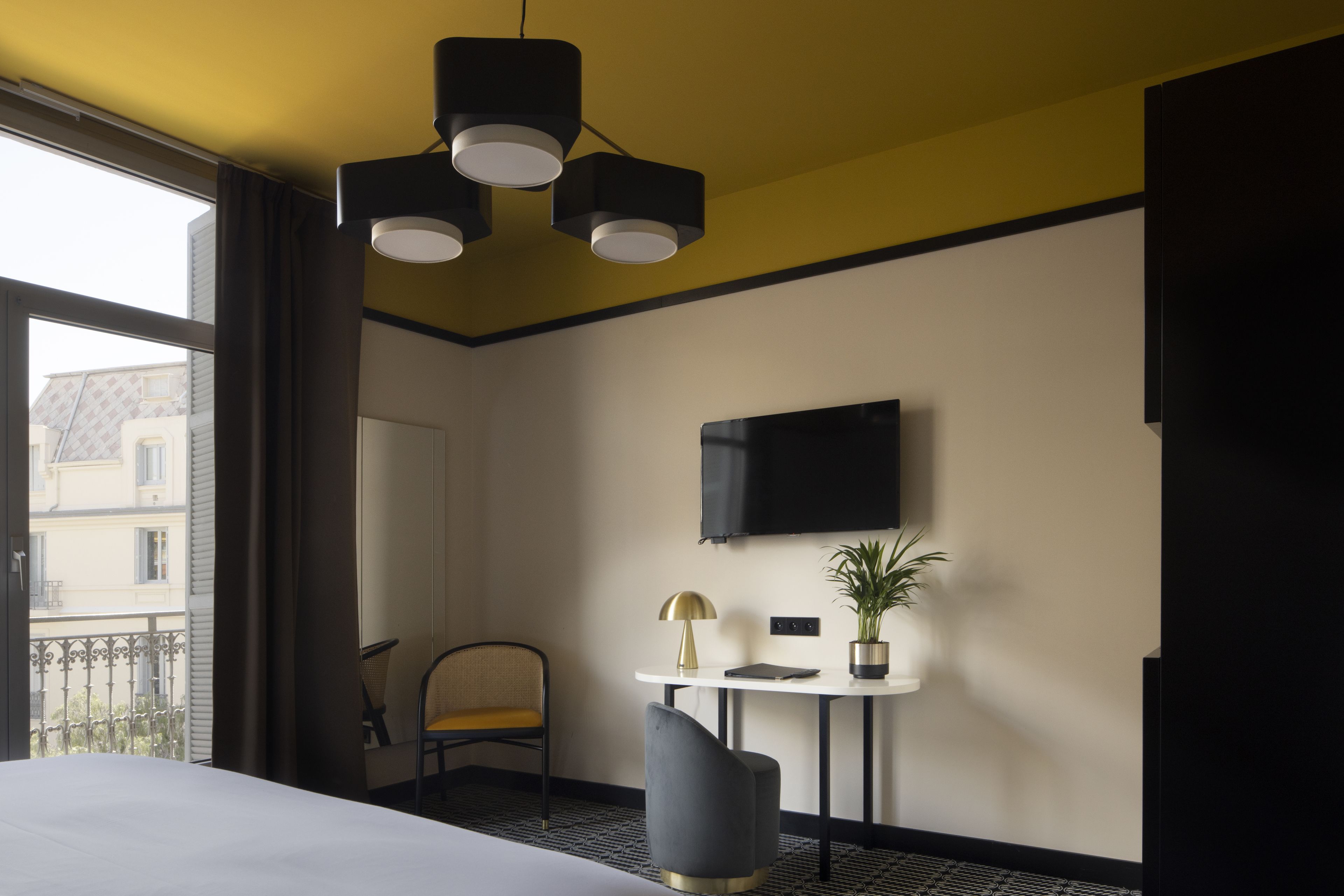 Quarto de hotel com cama, escrivaninha com luminária, poltrona, planta em vaso e televisão na parede.