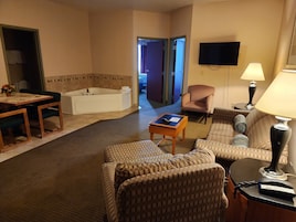 Quarto de hotel com área de estar, mesa de jantar, sofá, poltrona, televisão, abajur e banheira.
