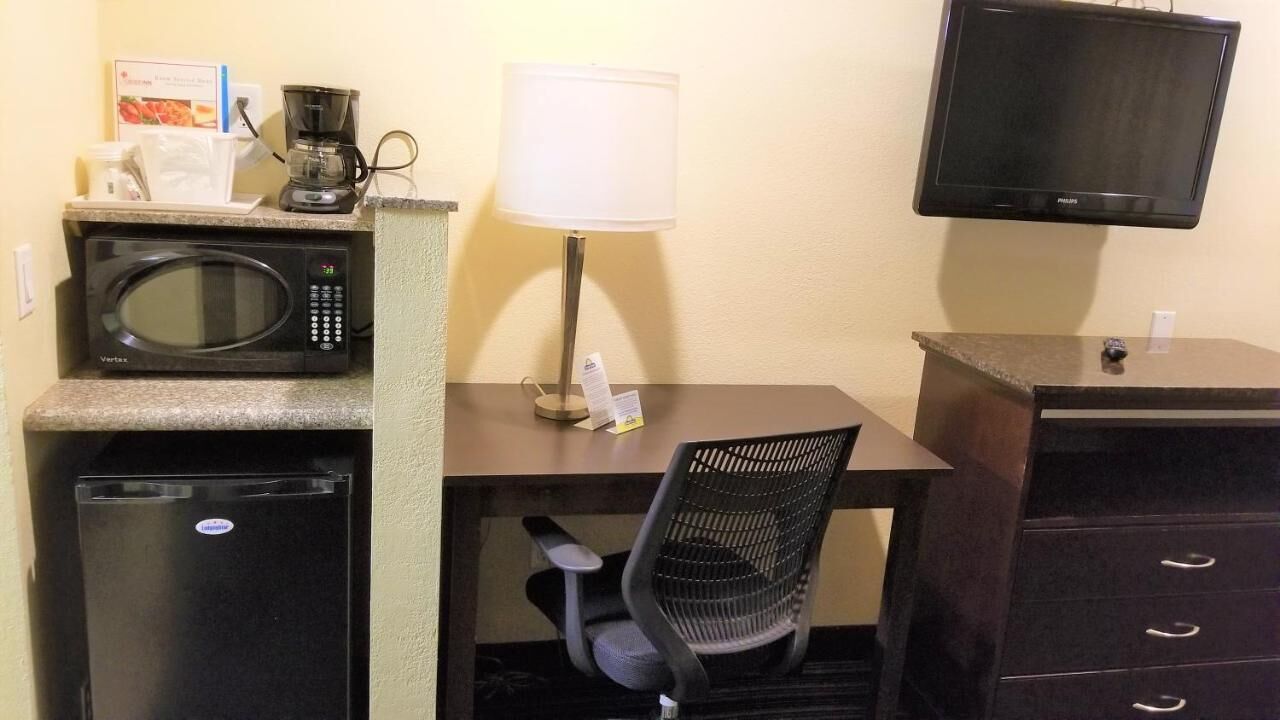 Quarto de hotel compacto com cozinha americana equipada com micro-ondas, cafeteira e geladeira. Possui uma mesa com cadeira e televisão sobre suporte.