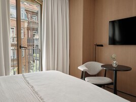 Quarto de hotel com uma cama grande, uma cadeira, uma mesinha, uma televisão e vista para a cidade pela janela.