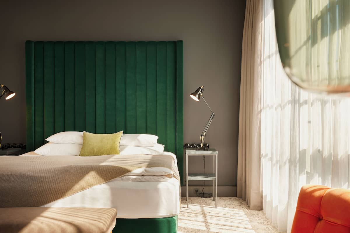 Quarto de hotel com uma cama grande, mesa de cabeceira com luminárias, uma cadeira e uma janela com cortinas.