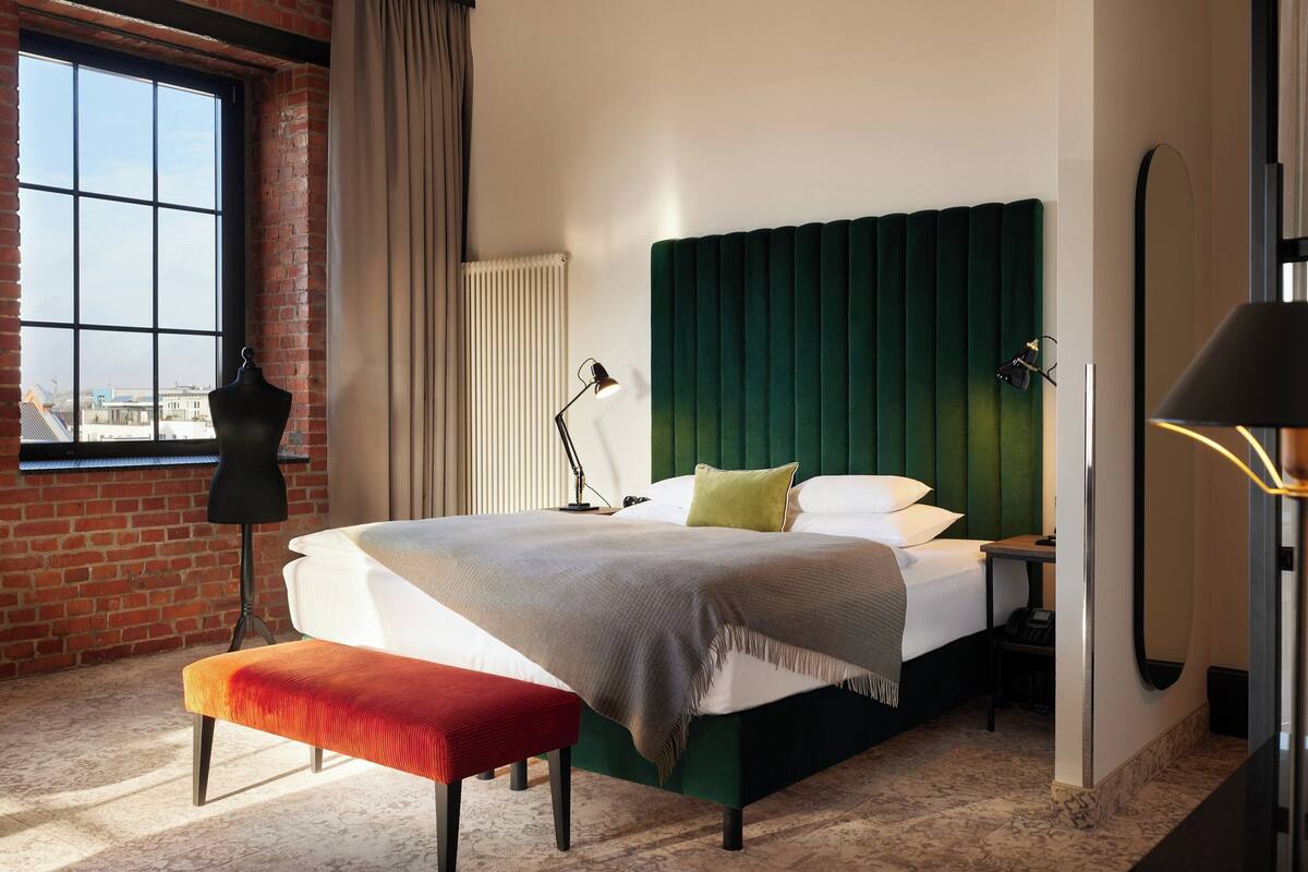 Quarto de hotel com uma janela grande, um manequim, uma cama com cabeceira verde, um banco e uma mesa com telefone.