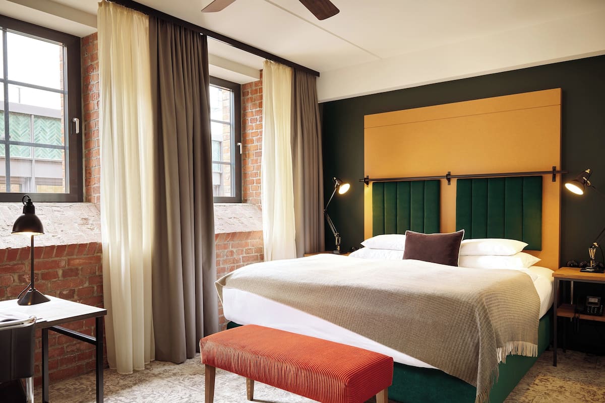 Quarto de hotel com uma cama grande, um banco, uma escrivaninha e uma janela com cortinas.
