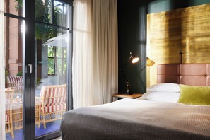 Quarto de hotel com cama, mesa de cabeceira, luminária, janela com cortinas, uma cadeira e vista para o exterior.