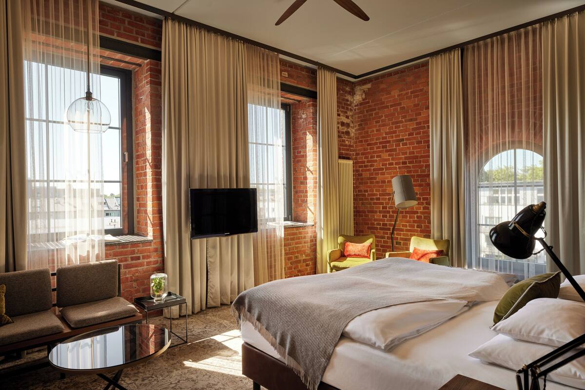 Quarto de hotel com parede de tijolos, uma cama, televisão, escrivaninha e uma cadeira.